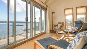 Quarto espaçoso com grandes portas de vidro deslizantes que dão para uma varanda com vista para o mar. Há duas cadeiras de descanso e uma cadeira de vime com almofadas.
