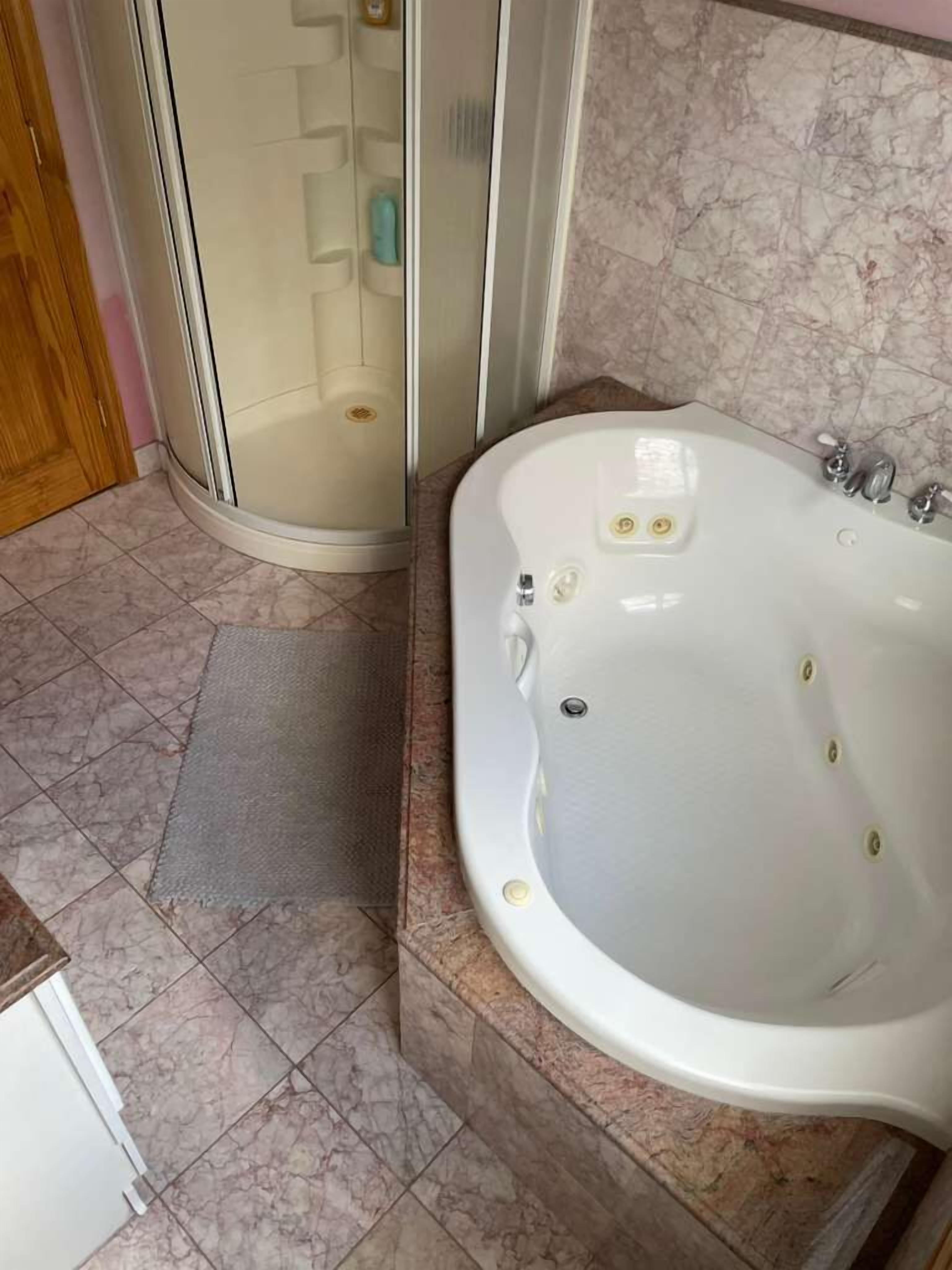 Um banheiro com piso de mármore, box de vidro e banheira branca com várias torneiras e painel de controle.