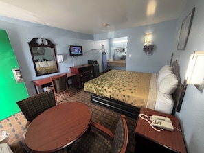 Quarto de hotel com cama, escrivaninha, cadeira, mesa, televisão, telefone e espelho.