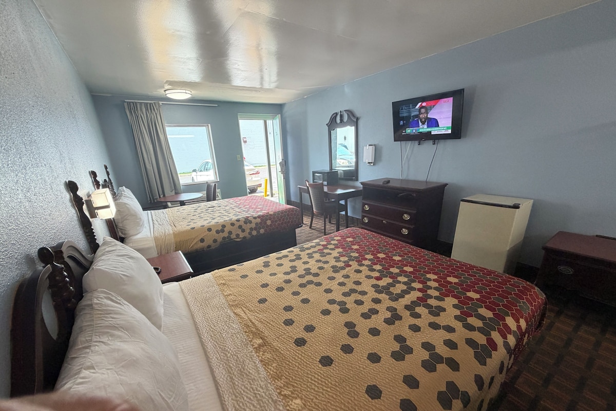 Quarto de hotel com duas camas, uma escrivaninha, uma cadeira, uma televisão e uma janela.