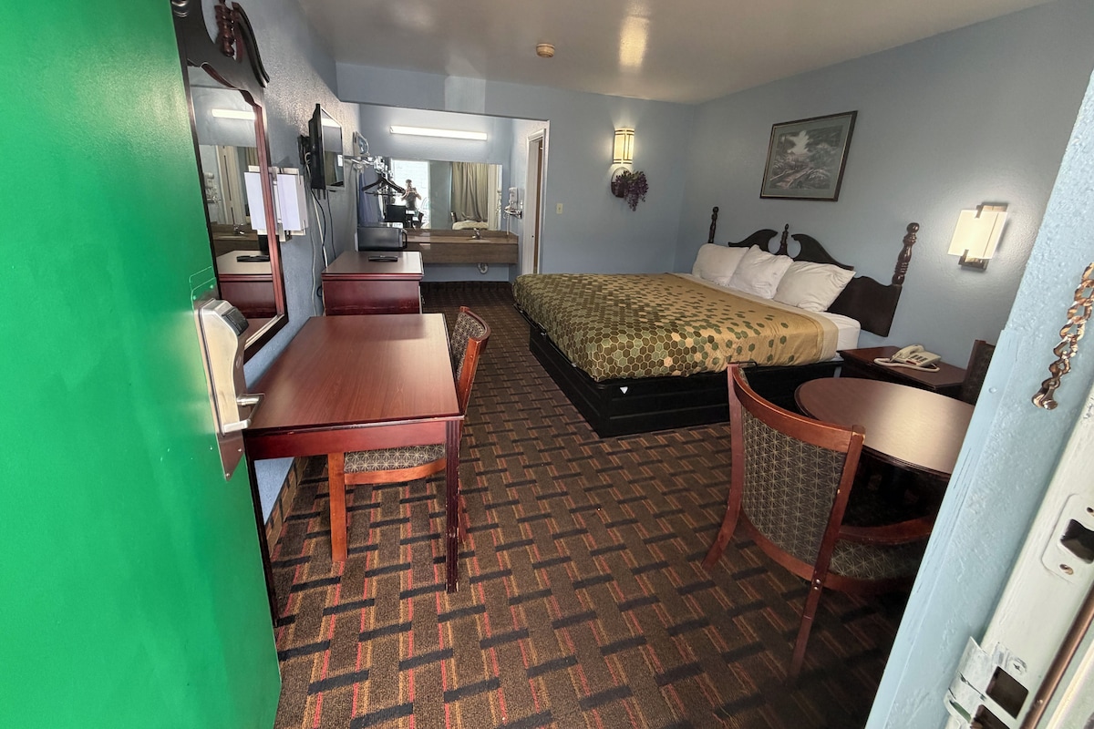 Quarto de hotel com cama, escrivaninha, cadeira, mesinha, espelho, abajur e um quadro na parede.
