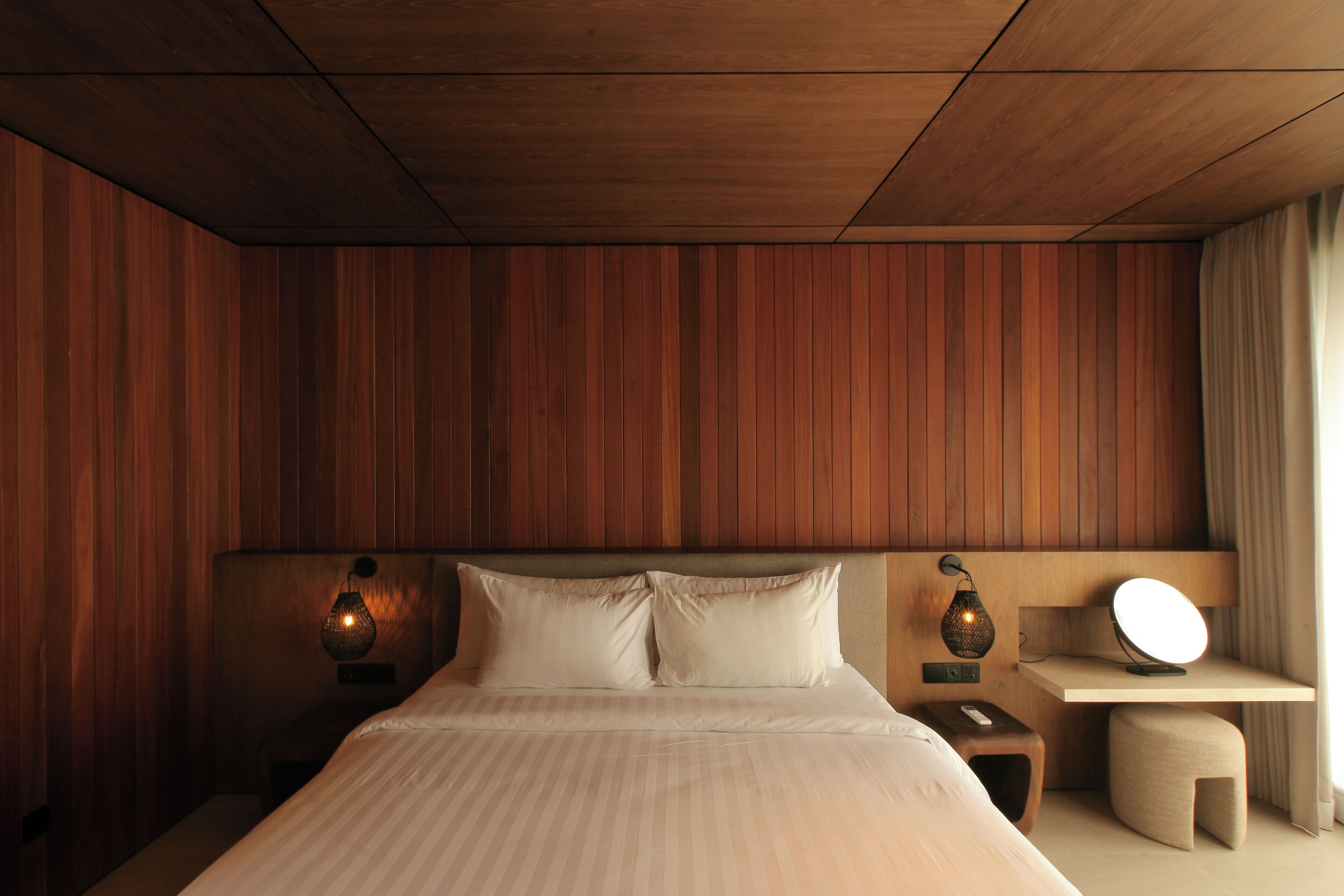 Quarto com cabeceira em painel de madeira, cama com roupa de cama branca, abajures, mesa de cabeceira e mesa lateral com luminária.