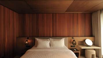 Quarto com cabeceira em painel de madeira, cama com roupa de cama branca, abajures, mesa de cabeceira e mesa lateral com luminária.