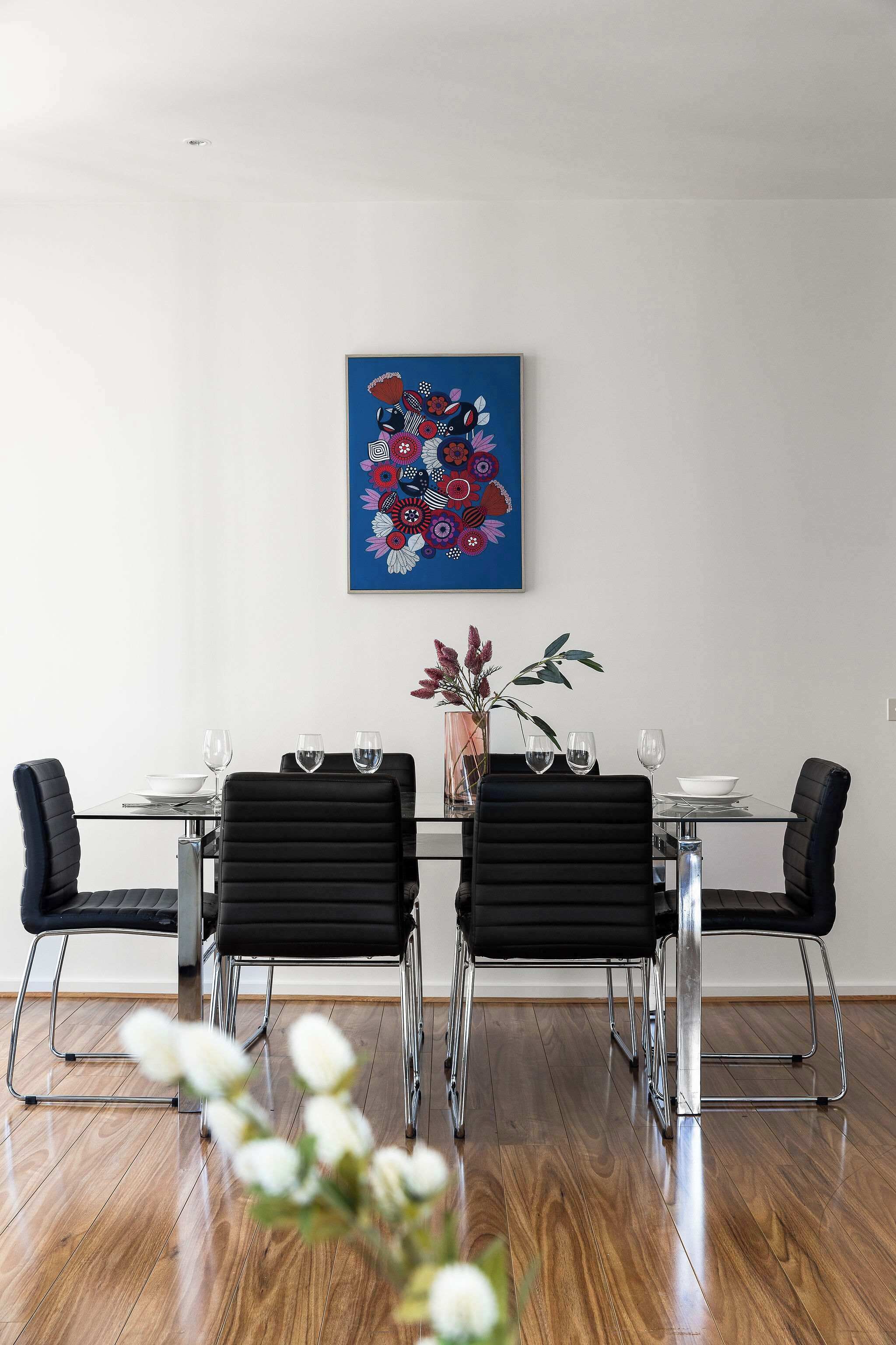 Uma sala de jantar com uma mesa de vidro, cadeiras pretas e um quadro com flores na parede.