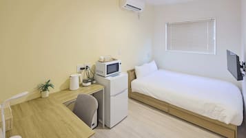 Quarto de hotel com cama, escrivaninha com computador, pequena geladeira, planta em vaso e ar-condicionado.