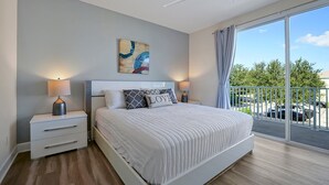 Quarto moderno com uma cama grande, duas mesas de cabeceira com luminárias, um ventilador de teto, uma janela ampla com vista para o exterior e uma obra de arte abstrata na parede.