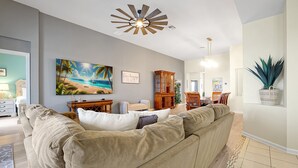 Uma sala de estar com sofá bege, ventilador de teto, pintura de uma praia e um armário de madeira.
