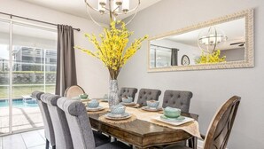 Uma sala de jantar com uma mesa grande, cadeiras estofadas cinzas, um vaso com flores amarelas e um espelho na parede.