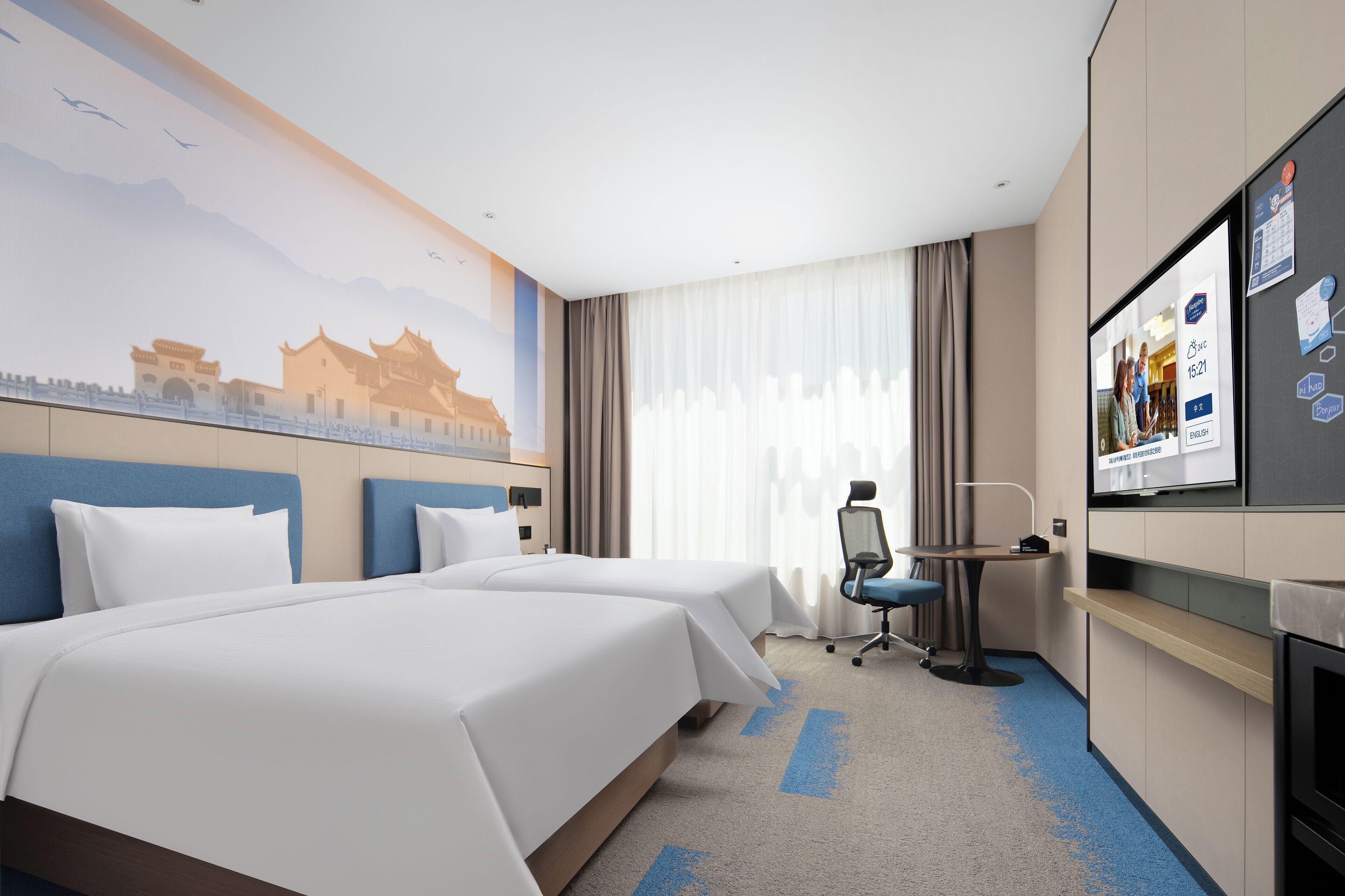 Quarto de hotel com duas camas, uma escrivaninha, uma TV e um mural de um edifício tradicional.