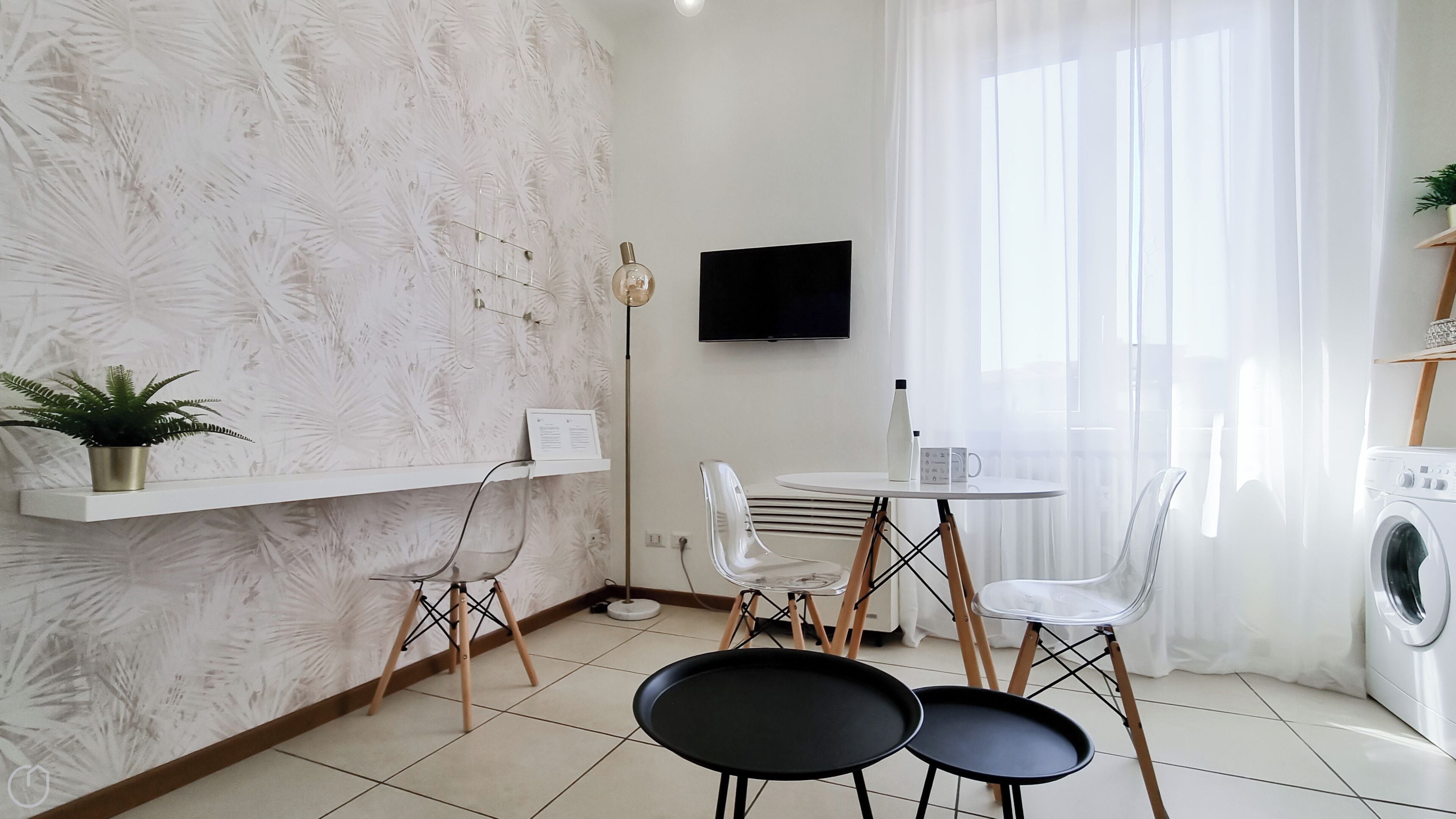Uma sala de estar moderna com parede de padrão branco, uma TV de tela plana, uma mesa de jantar redonda com cadeiras, uma pequena planta em vaso e uma máquina de lavar roupa.