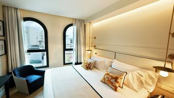 Quarto de hotel com uma cama grande, uma cadeira, uma escrivaninha e vista da cidade através de janelas em arco.
