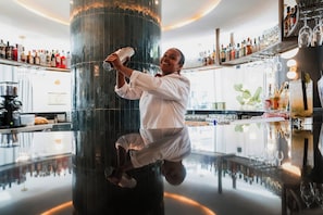 Uma área de bar com piso polido, um bartender preparando uma bebida, e prateleiras repletas de garrafas e copos.