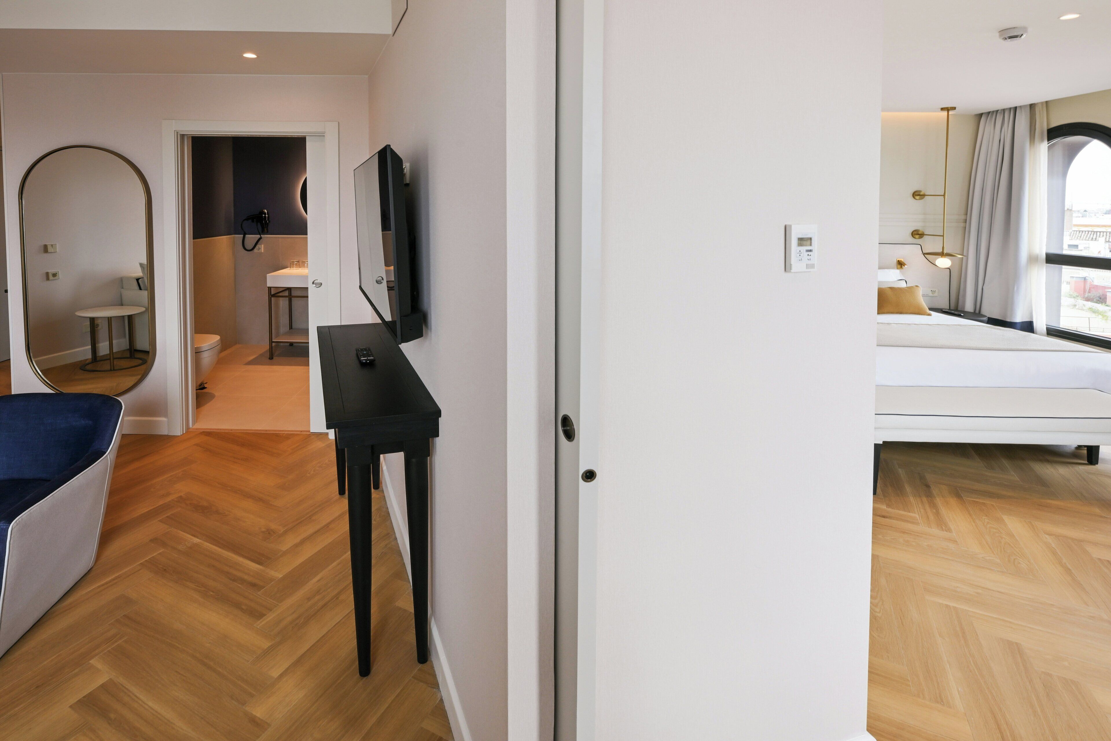 Quarto de hotel com piso de madeira, uma cama, uma escrivaninha com cadeira, um espelho, uma televisão e um banheiro com vaso sanitário e pia.