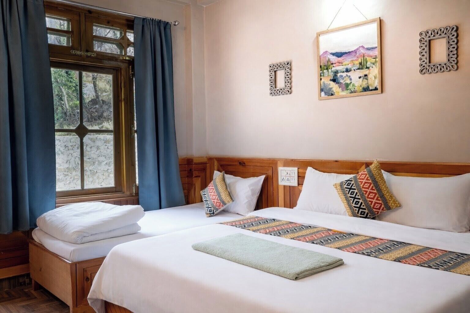 Uma cama de casal com roupa de cama branca, cabeceira de madeira e almofadas estampadas. Há dois interruptores de luz fixados na parede e um quadro emoldurado acima da cama.