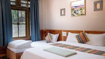 Uma cama de casal com roupa de cama branca, cabeceira de madeira e almofadas estampadas. Há dois interruptores de luz fixados na parede e um quadro emoldurado acima da cama.
