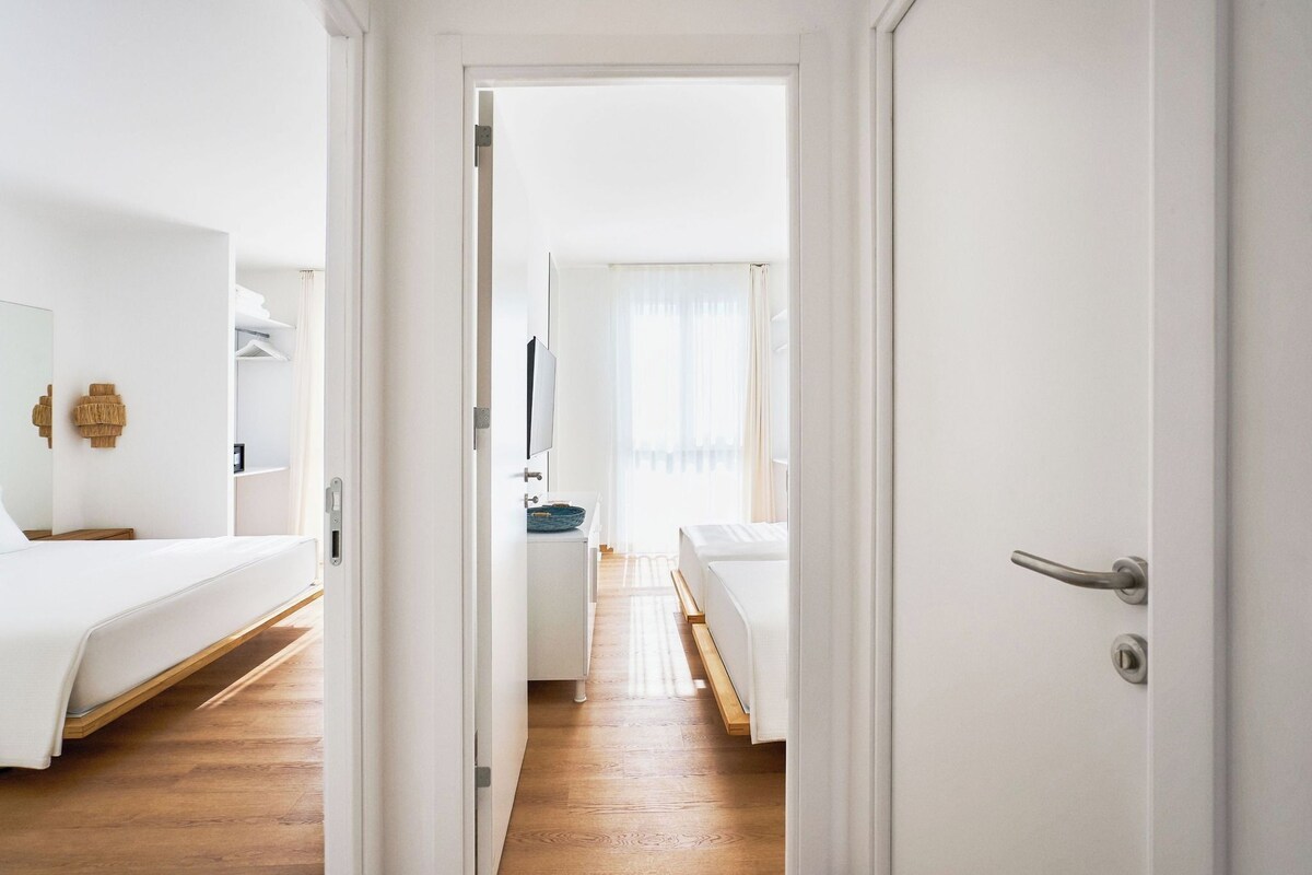 Quarto de hotel com piso de madeira, uma cama, uma mesinha de cabeceira e um banheiro visível por uma porta aberta.