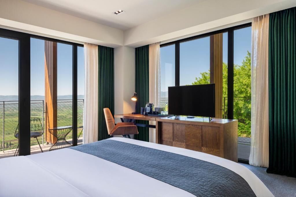 Quarto de hotel moderno com uma cama grande, uma escrivaninha com televisão de tela plana e uma varanda com vista para o campo.