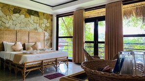 Quarto com parede de pedra, cama com almofadas, cadeira de vime e janela com cortinas.