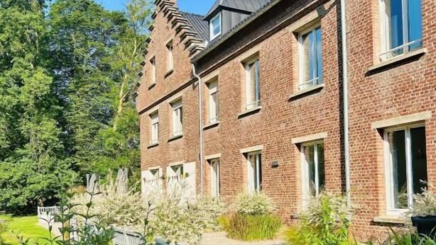 Um edifício de tijolos com várias janelas e telhado inclinado, rodeado por vegetação e um banco.