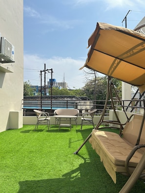 Terraço no topo do prédio com grama sintética, cadeira de balanço, uma mesa e vista para a paisagem urbana.