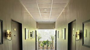 Corredor comprido e estreito, com portas de ambos os lados, plantas em vasos e teto com iluminação embutida.