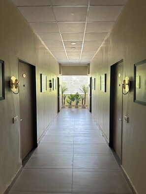 Corredor comprido e estreito, com portas de ambos os lados, plantas em vasos e teto com iluminação embutida.