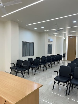 Sala de conferência com fileiras de cadeiras, uma mesa de madeira, luminárias no teto e um aparelho de ar condicionado.