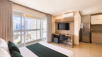Quarto de hotel com cama, escrivaninha, televisão e cozinha compacta.