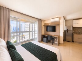 Quarto de hotel com cama, escrivaninha, televisão e cozinha compacta.