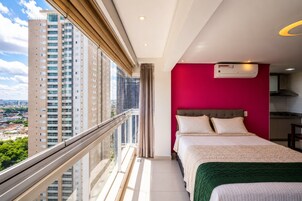Quarto moderno com cama grande, parede de destaque vermelha e varanda com vista para a cidade.