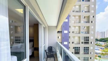 Varanda com vista para um edifício alto, uma cadeira e uma cama com janela.