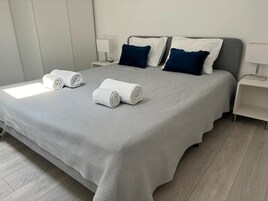 Cama bem arrumada com colcha cinza, duas toalhas brancas enroladas, dois travesseiros azul escuro e uma mesa de cabeceira branca com abajur.