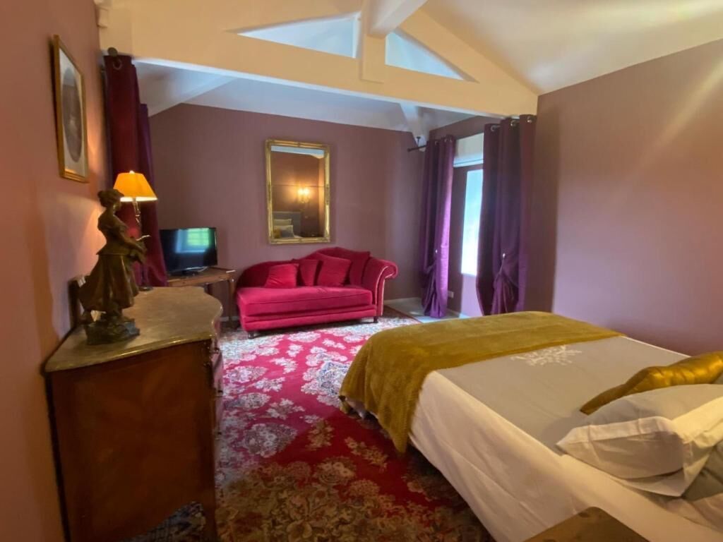 Quarto de hotel com cama, sofá vermelho, escrivaninha com luminária, televisão e janela com cortinas.