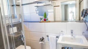 Uma casa de banho moderna com cabine de duche em vidro, sanita branca, lavatório com espelho e uma pequena estante com plantas.