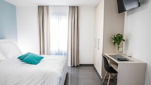 Quarto de hotel com cama, escrivaninha, cadeira, televisão e janela com cortinas.