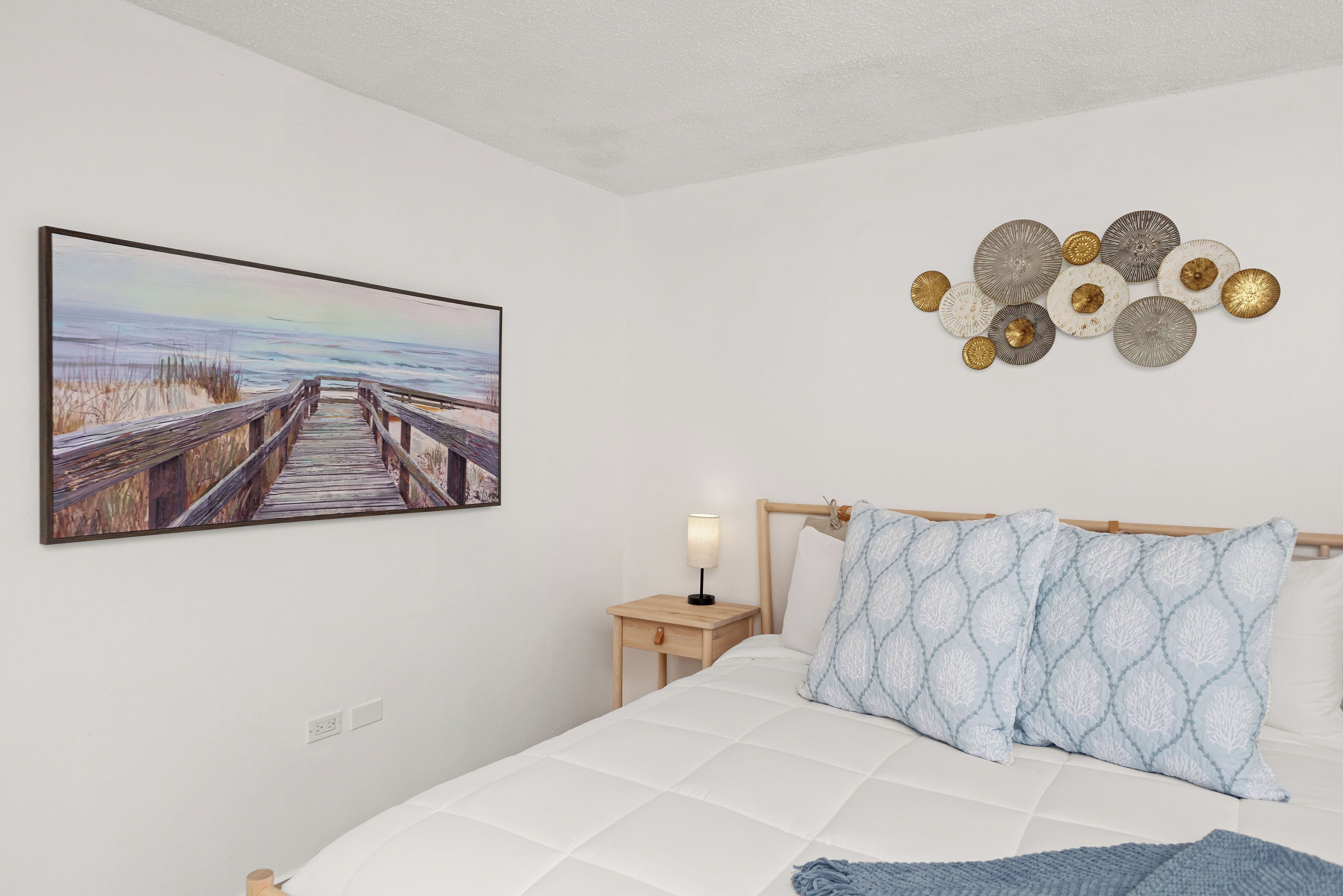 Quarto com cama, mesa de cabeceira com abajur, quadro de praia na parede e decoração de parede.