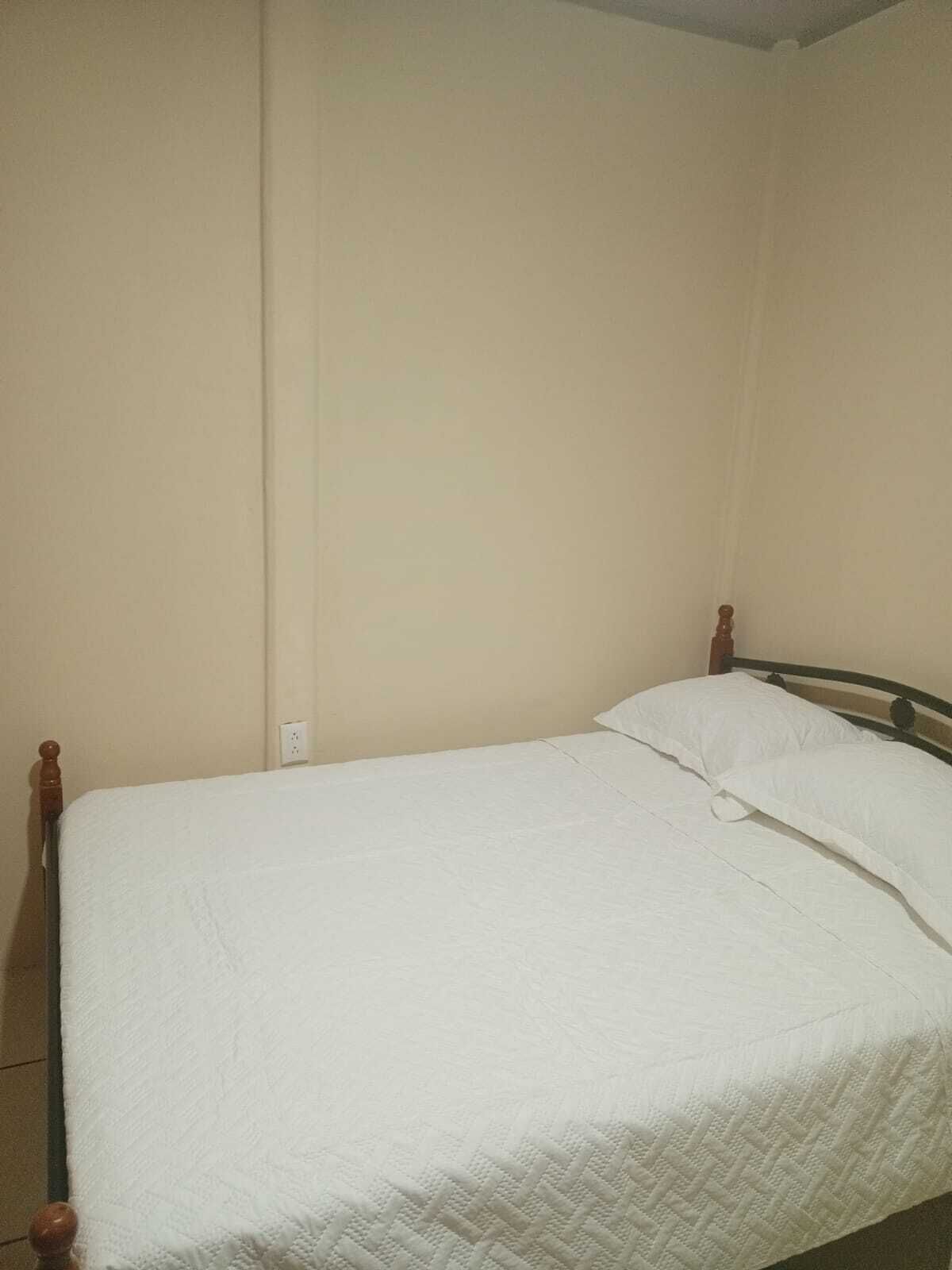 Uma cama de solteiro com estrutura de madeira e roupa de cama branca num quarto com paredes lisas e uma tomada elétrica visível.