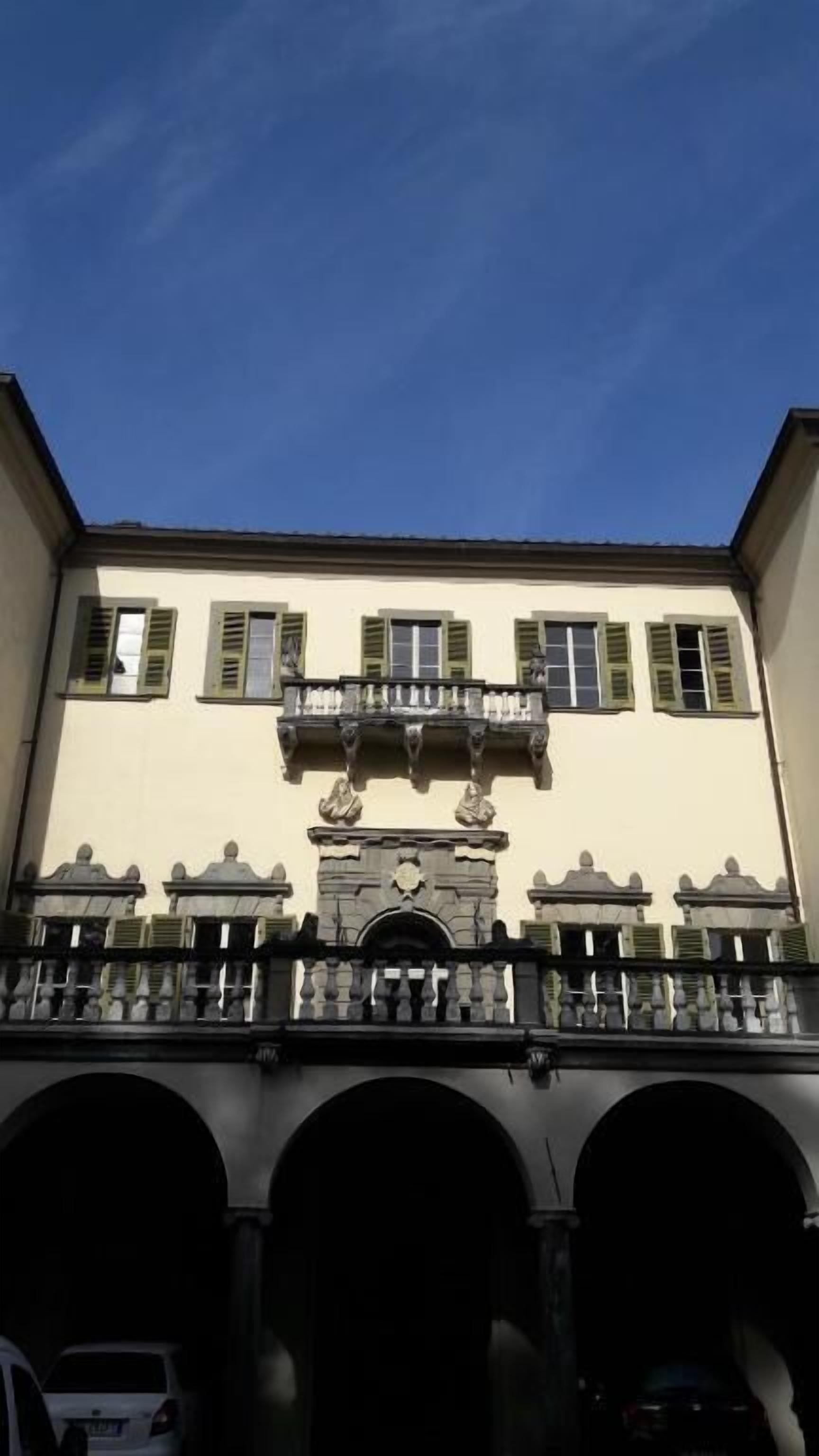 Um edifício com varanda, entrada central com arco decorativo e uma série de janelas com persianas.