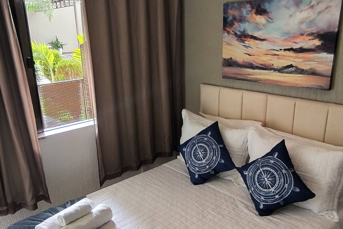 Quarto com cama, almofadas, cobertor, pintura e vista para o verde lá fora.