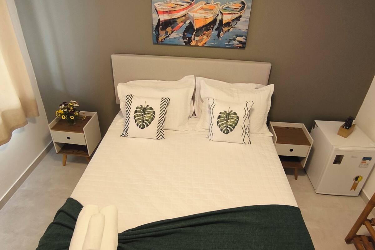 Cama bem arrumada com manta verde, almofadas brancas e quadro de barcos na parede.