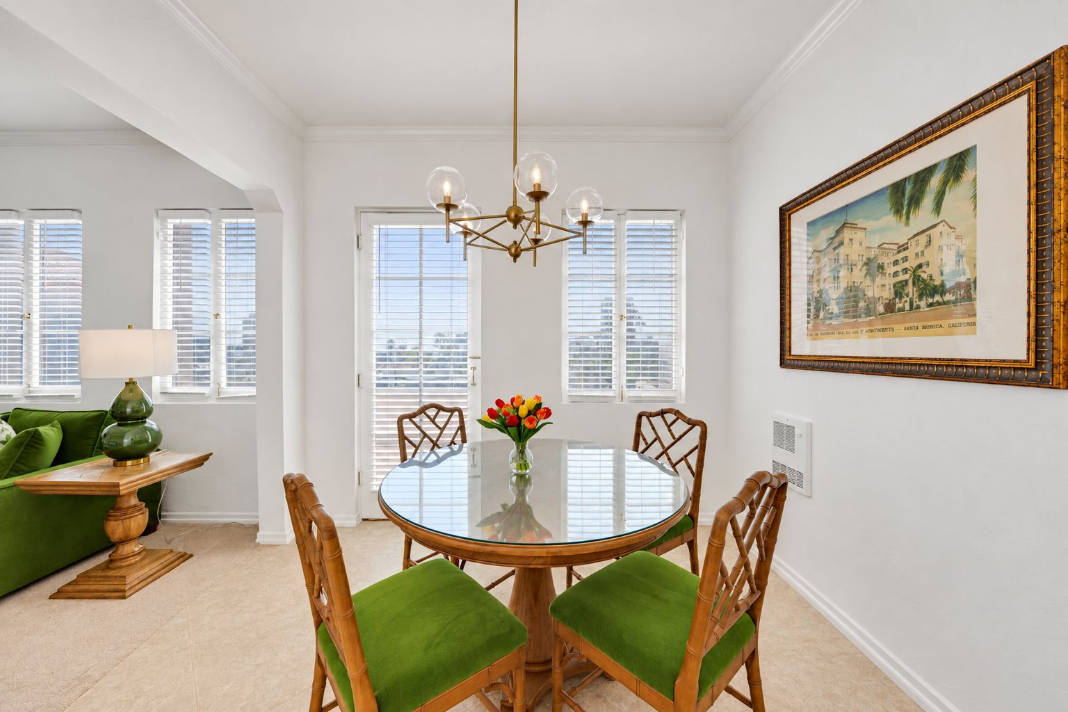 Uma sala de jantar com uma mesa de vidro redonda, cadeiras estofadas em verde, um candelabro e um quadro na parede.