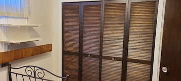 Quarto com guarda-roupa de madeira, estrutura de cama de metal e parede branca com prateleira.
