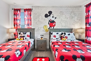 Quarto com duas camas, cada uma com roupa de cama temática do Mickey Mouse. Há adesivos de parede, cortinas e almofadas com o tema do Mickey Mouse.