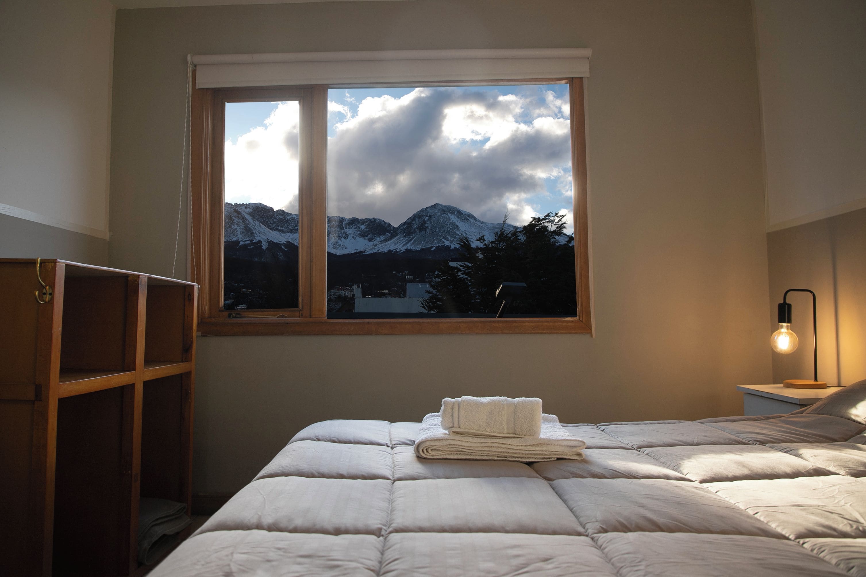 Quarto com cama, janela com vista para as montanhas, mesa de cabeceira com abajur e guarda-roupa de madeira.