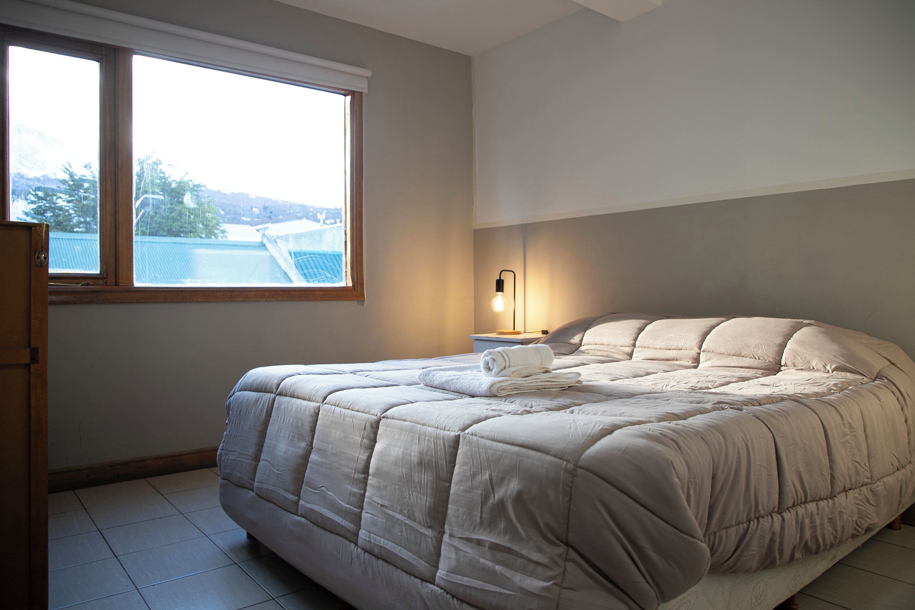 Quarto com uma cama grande, mesa de cabeceira com abajur e janela com vista para edifícios.
