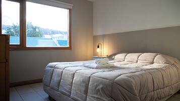 Quarto com uma cama grande, mesa de cabeceira com abajur e janela com vista para edifícios.
