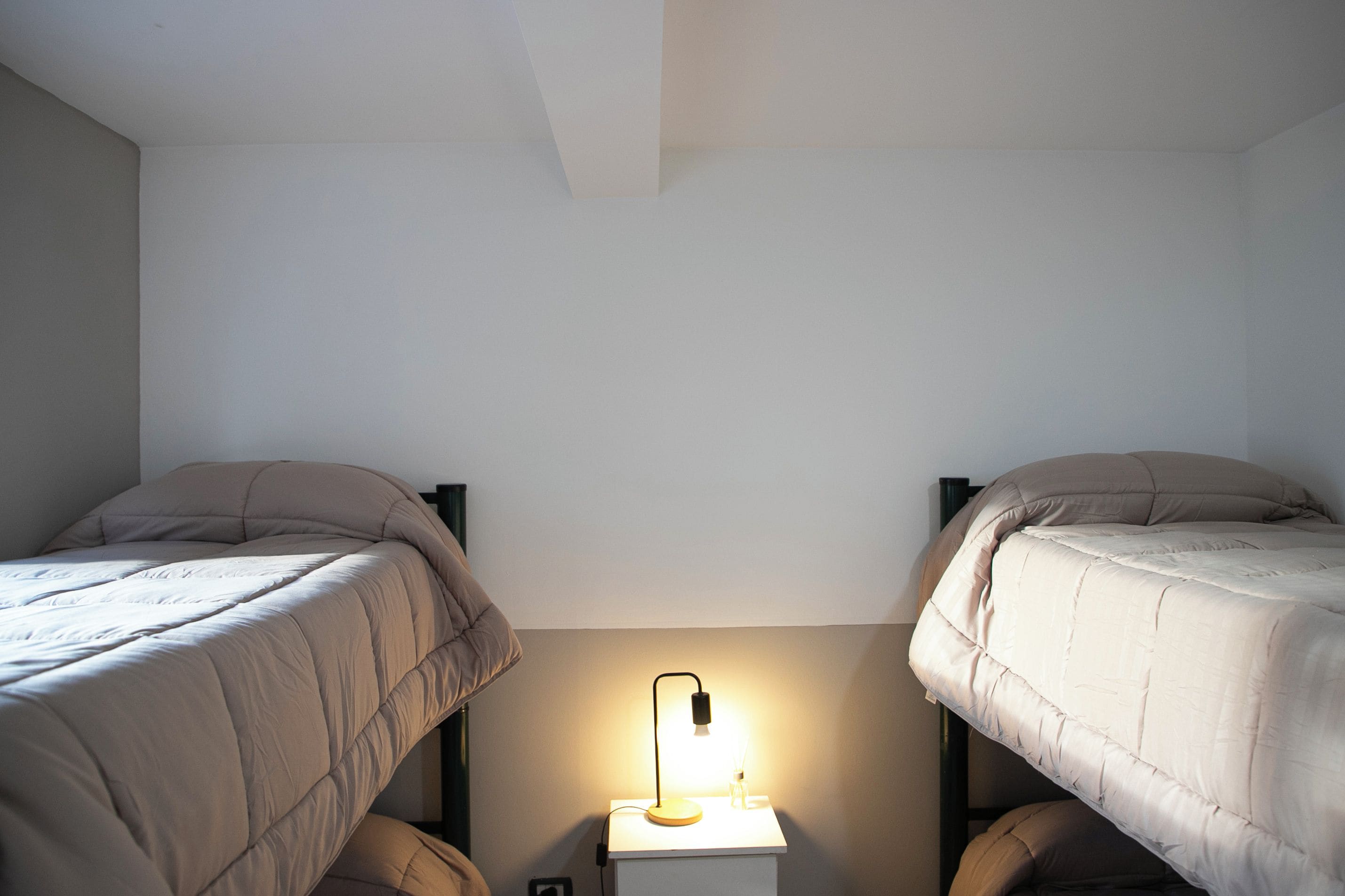 Quarto duplo com duas camas de solteiro, uma mesa de cabeceira com abajur e uma parede branca.