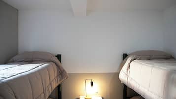 Quarto duplo com duas camas de solteiro, uma mesa de cabeceira com abajur e uma parede branca.