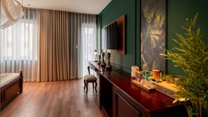 Quarto de hotel com piso de madeira, uma cama, uma escrivaninha com televisão de tela plana, uma cadeira e uma janela grande com cortinas.
