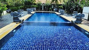 Uma piscina retangular com piso em azulejo, rodeada por espreguiçadeiras e palmeiras.